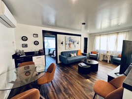 Uma sala de estar moderna com sofá, cadeiras, mesa de centro e televisão. Há uma área de jantar com mesa de vidro e cadeiras. O cômodo possui piso de madeira e uma grande janela com cortinas.