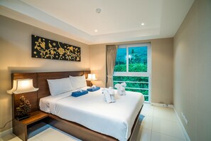Quarto de hotel com uma cama grande, duas luminárias de cabeceira, um painel decorativo na parede, uma janela com vista para o verde e uma pequena mesa com abajur.