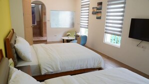 Quarto de hotel com duas camas, uma mesa pequena com uma cadeira, televisão e janela com persianas.