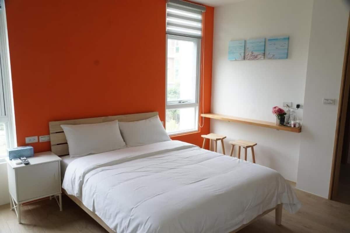 Quarto com cama, mesa de cabeceira, janela e prateleira com objetos.
