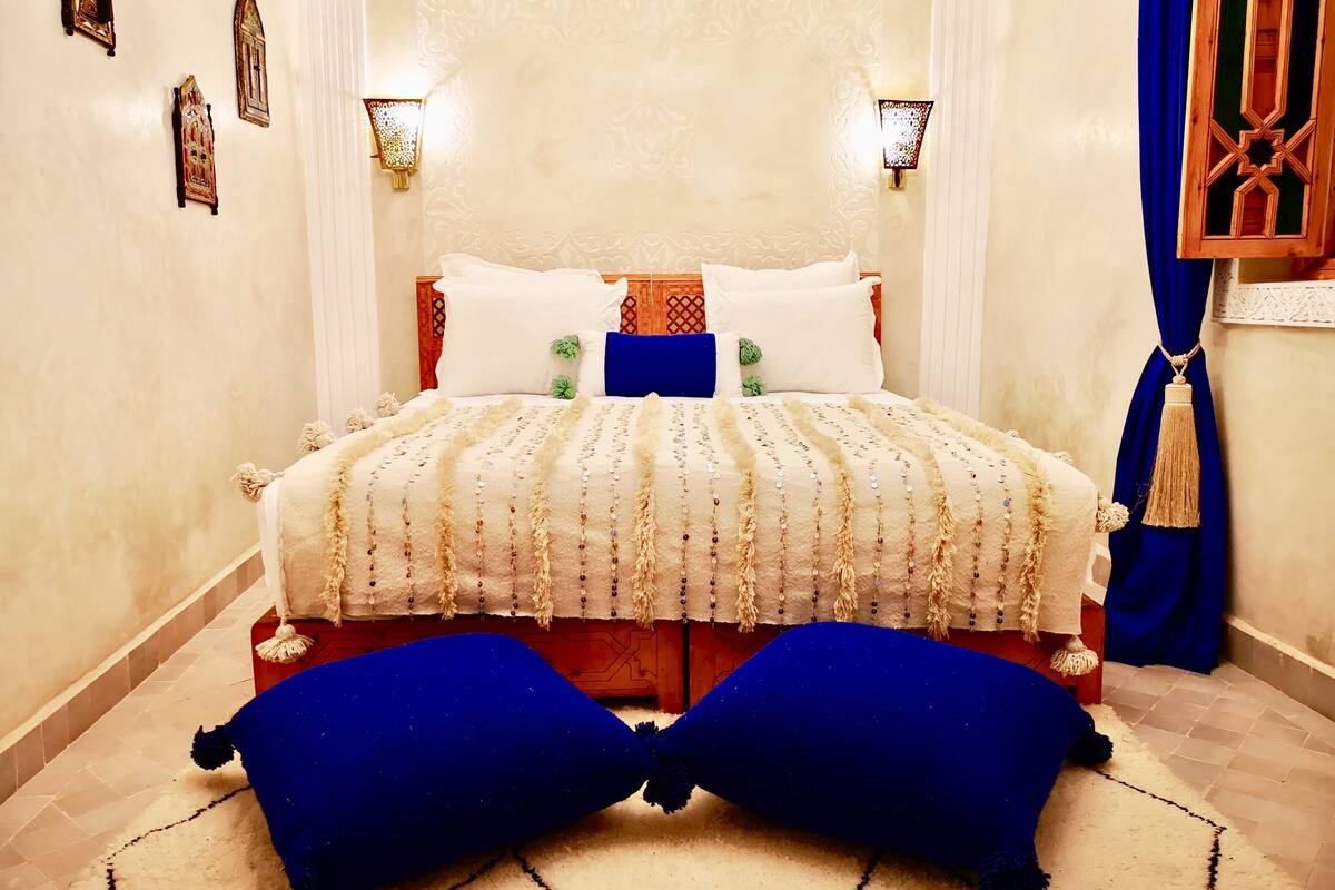 Uma cama com cabeceira decorativa, duas luminárias na parede e uma cortina azul.