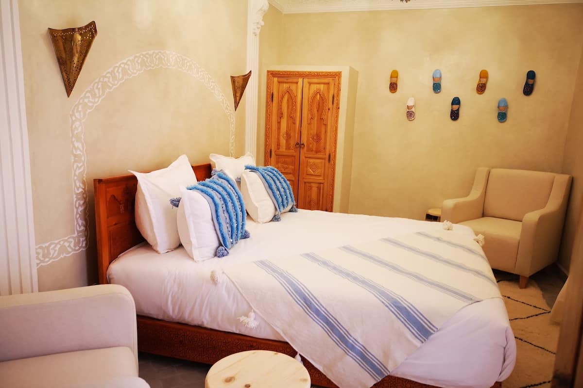 Quarto bem organizado com cama grande, guarda-roupa de madeira, um banquinho redondo pequeno e uma poltrona bege.