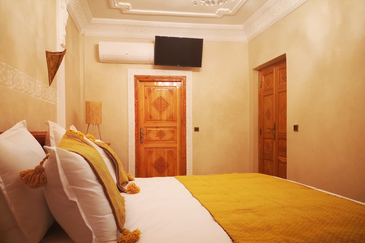 Quarto de hotel com cama, almofadas, cobertor, televisão e porta.