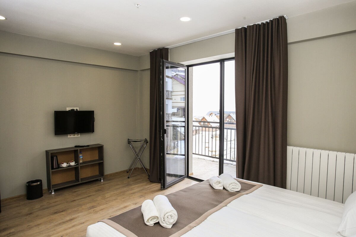 Quarto de hotel com cama, televisão, prateleira pequena com produtos de higiene e varanda com cortinas.