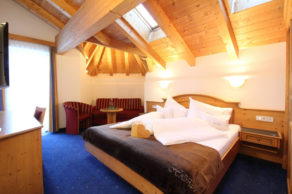 Quarto de hotel com uma cama grande, teto de madeira, sofá, mesa pequena e televisão.