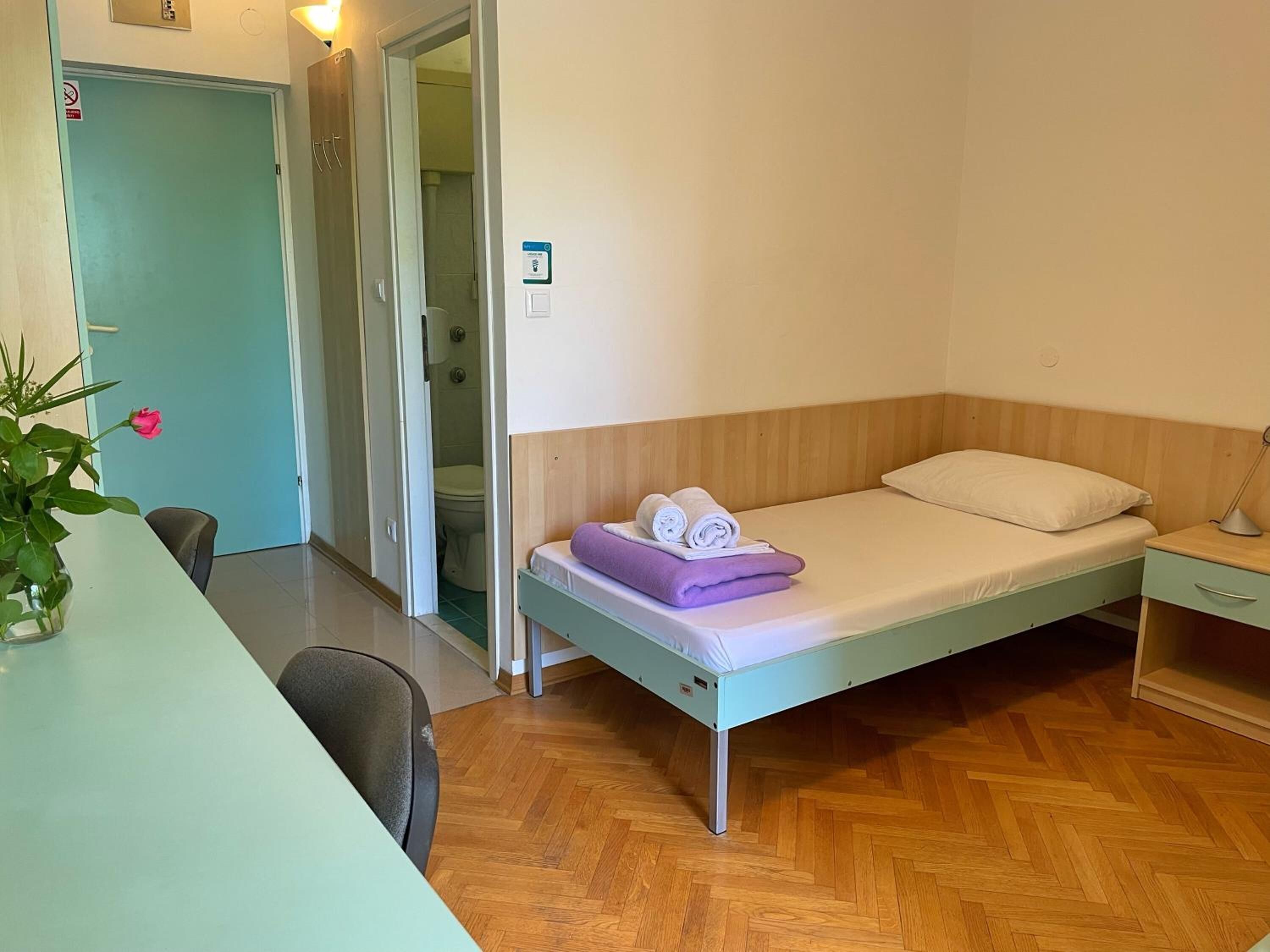 Um quarto pequeno e arrumado, com uma cama de solteiro, mesa de cabeceira, cadeira e uma mesa com um vaso de flores.