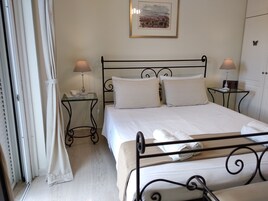 Cama bem arrumada com lençóis brancos, duas mesas de cabeceira com abajures, um quadro na parede e uma janela com cortinas.