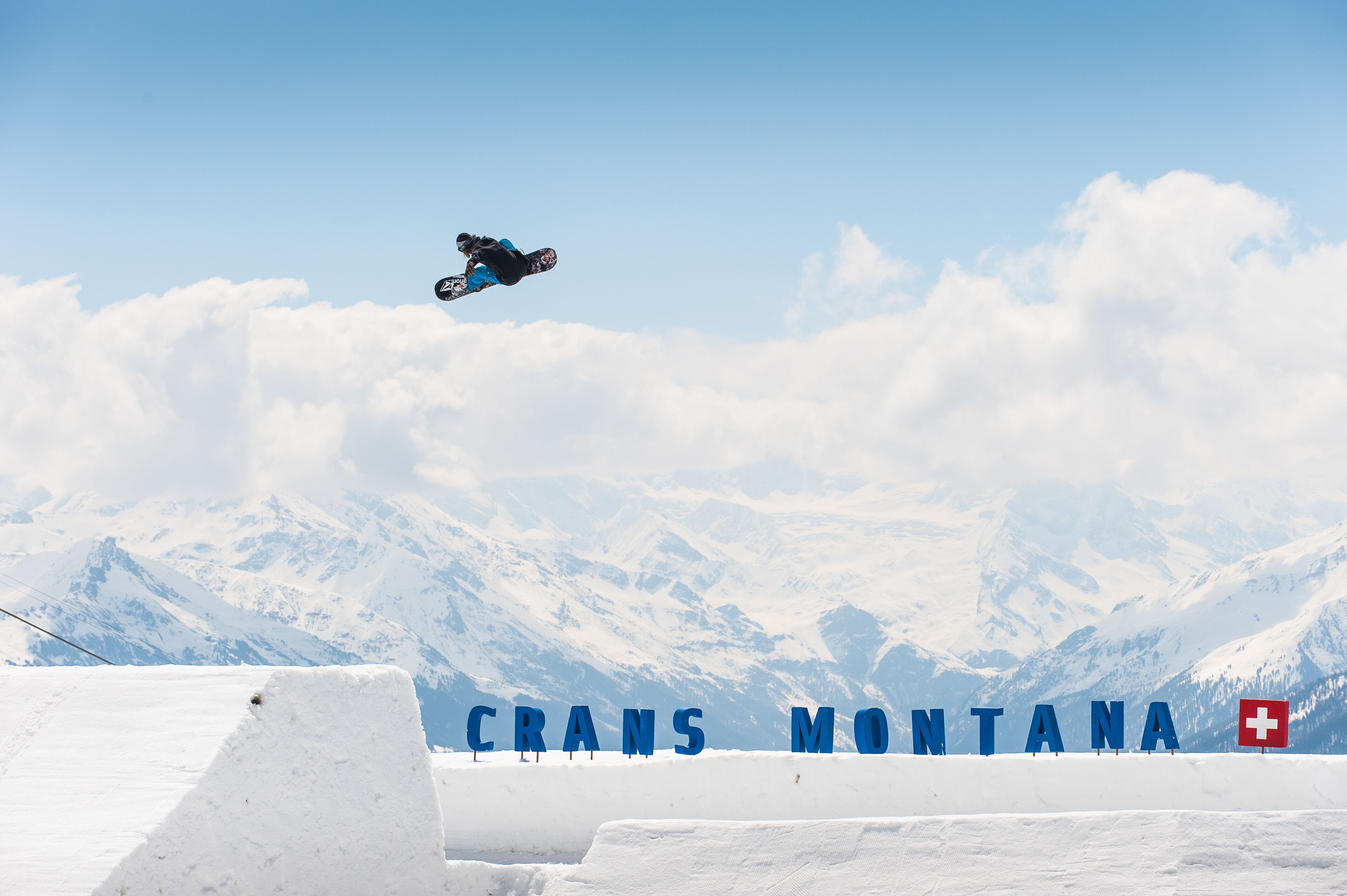 Um snowboarder no ar, com montanhas cobertas de neve e o texto "CRANS MONTANA" ao fundo.
