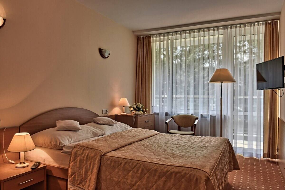 Quarto de hotel com uma cama grande, duas mesas de cabeceira com abajures, uma cadeira, uma televisão e uma janela com cortinas.