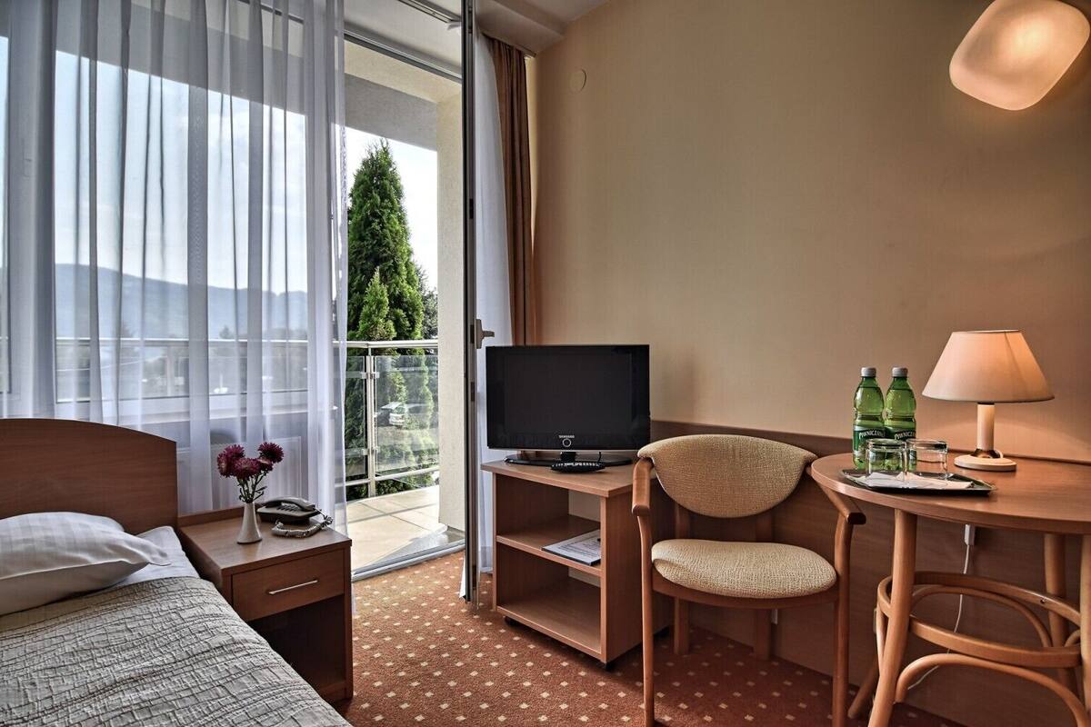 Quarto de hotel com cama, escrivaninha com televisão, cadeira, mesinha com bebidas e varanda com vista para árvores.