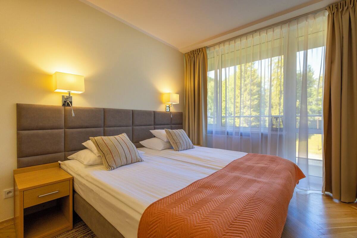Quarto de hotel com uma cama grande, duas luminárias de cabeceira, mesa de apoio e vista para o exterior através de cortinas translúcidas.