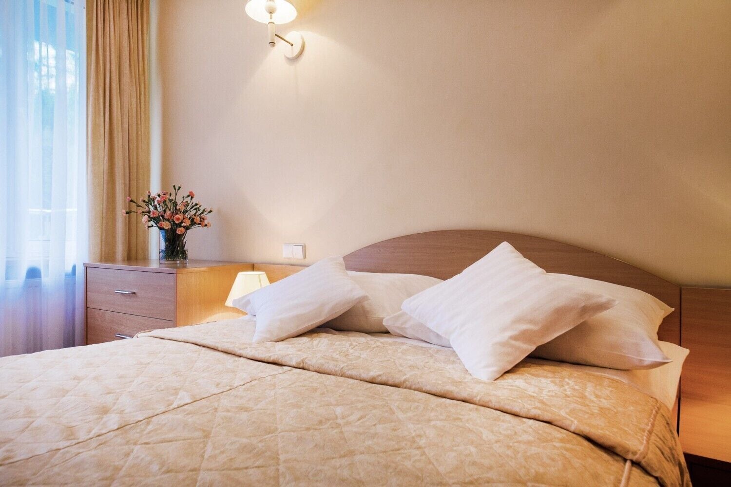 Uma cama bem arrumada com cobertura acolchoada e duas almofadas, um abajur, um vaso com flores e uma mesa de cabeceira de madeira.
