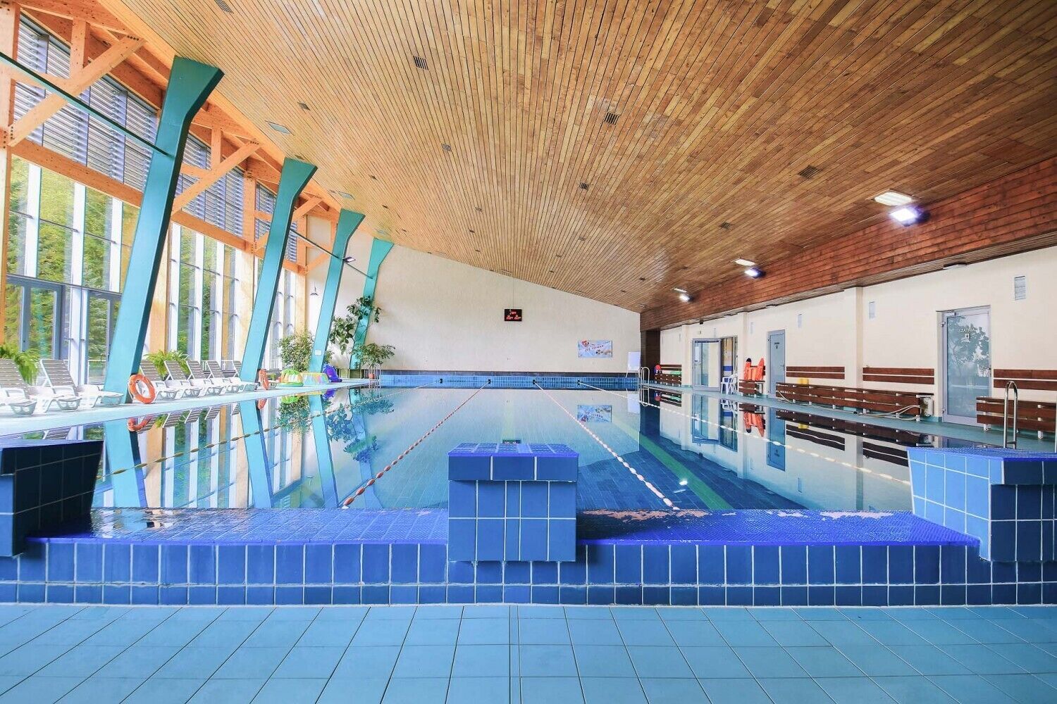 Piscina interna com teto de madeira, chão de azulejos azuis e janelas iluminadas pelo sol.