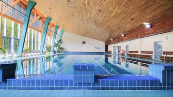 Piscina interna com teto de madeira, chão de azulejos azuis e janelas iluminadas pelo sol.