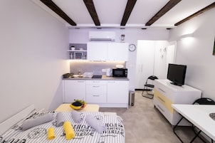 Quarto de hotel compacto com cozinha compacta, uma cama com roupa de cama listrada, uma mesa pequena com uma tigela de fruta e uma televisão em um suporte.