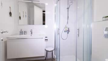 Um banheiro moderno com chuveiro em box de vidro, toucador branco com espelho e um banquinho pequeno.