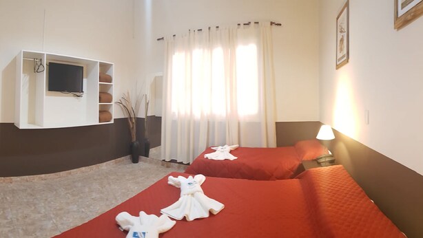 Quarto de hotel com duas camas, televisão na parede, tapete vermelho e abajur na mesinha de cabeceira.