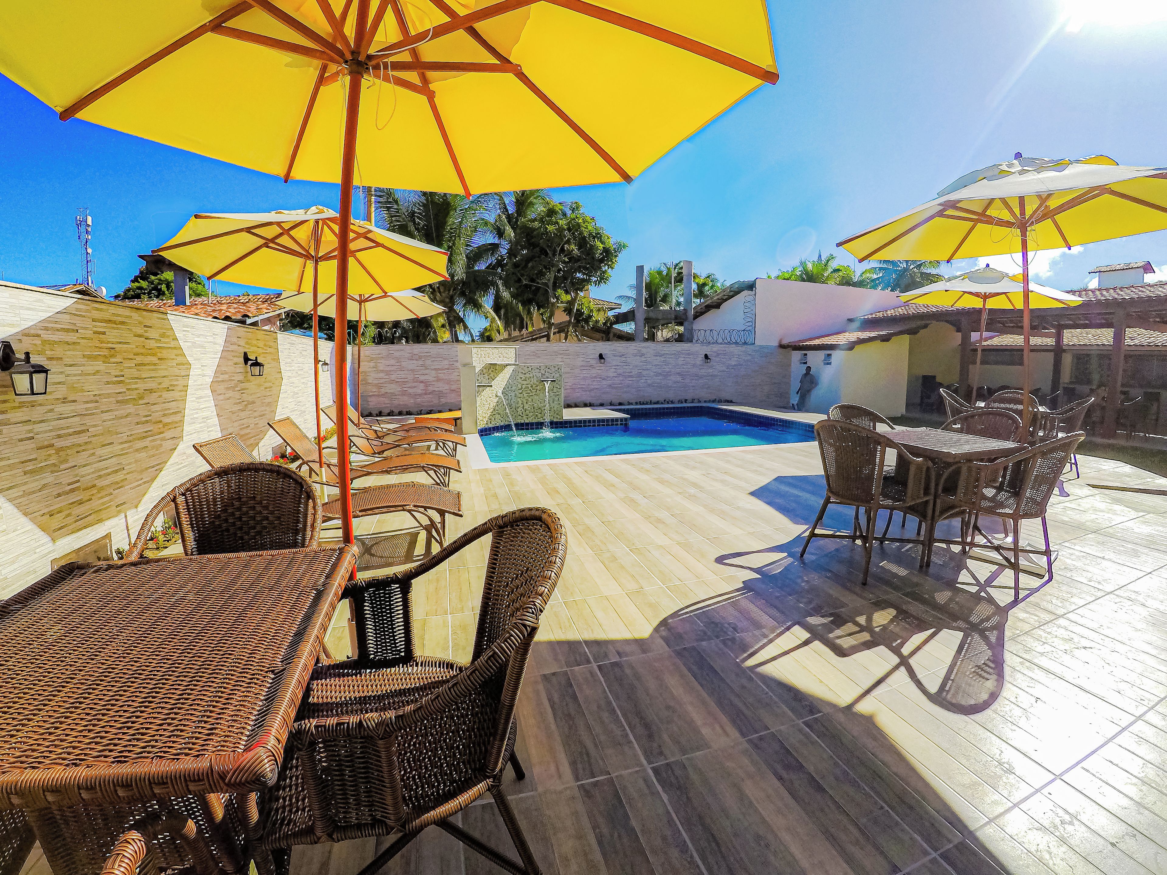 Uma área à beira da piscina com móveis de vime, guarda-sóis amarelos e piso de azulejos.