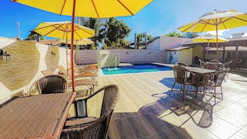 Uma área à beira da piscina com móveis de vime, guarda-sóis amarelos e piso de azulejos.