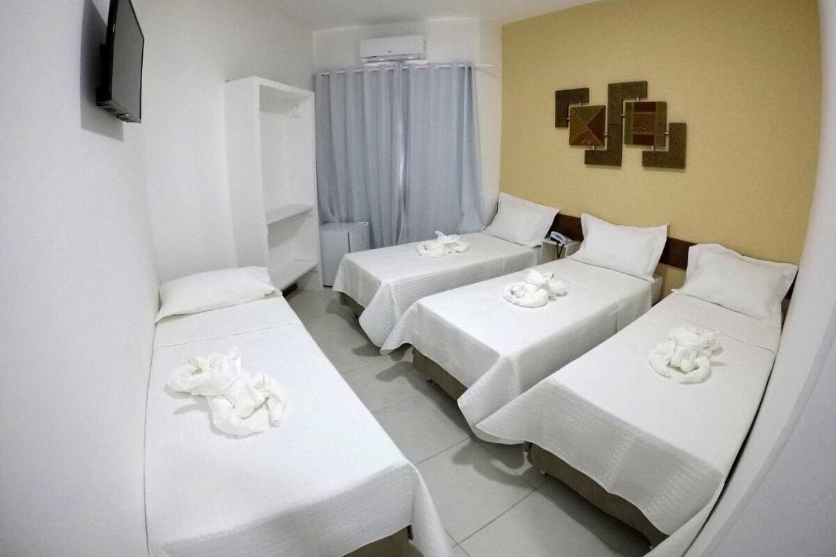 Quarto de hotel com três camas de solteiro, TV fixada na parede e janela com cortinas.