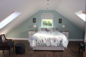 Quarto com teto inclinado, cama com roupa de cama florida, duas mesas de cabeceira com abajures, uma cadeira e uma janela com vista para o verde.