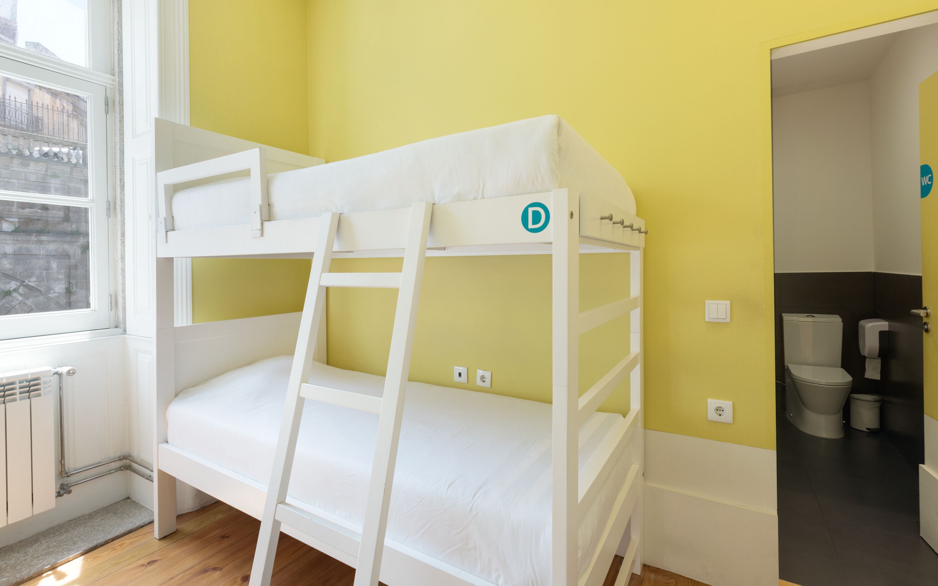 Uma cama beliche com escada, uma janela e vista para a rua.
