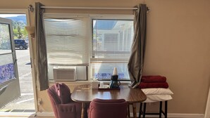 Quarto com uma mesa de madeira, duas cadeiras vermelhas, uma janela com persianas, uma toalha vermelha e vista para um estacionamento e montanhas.