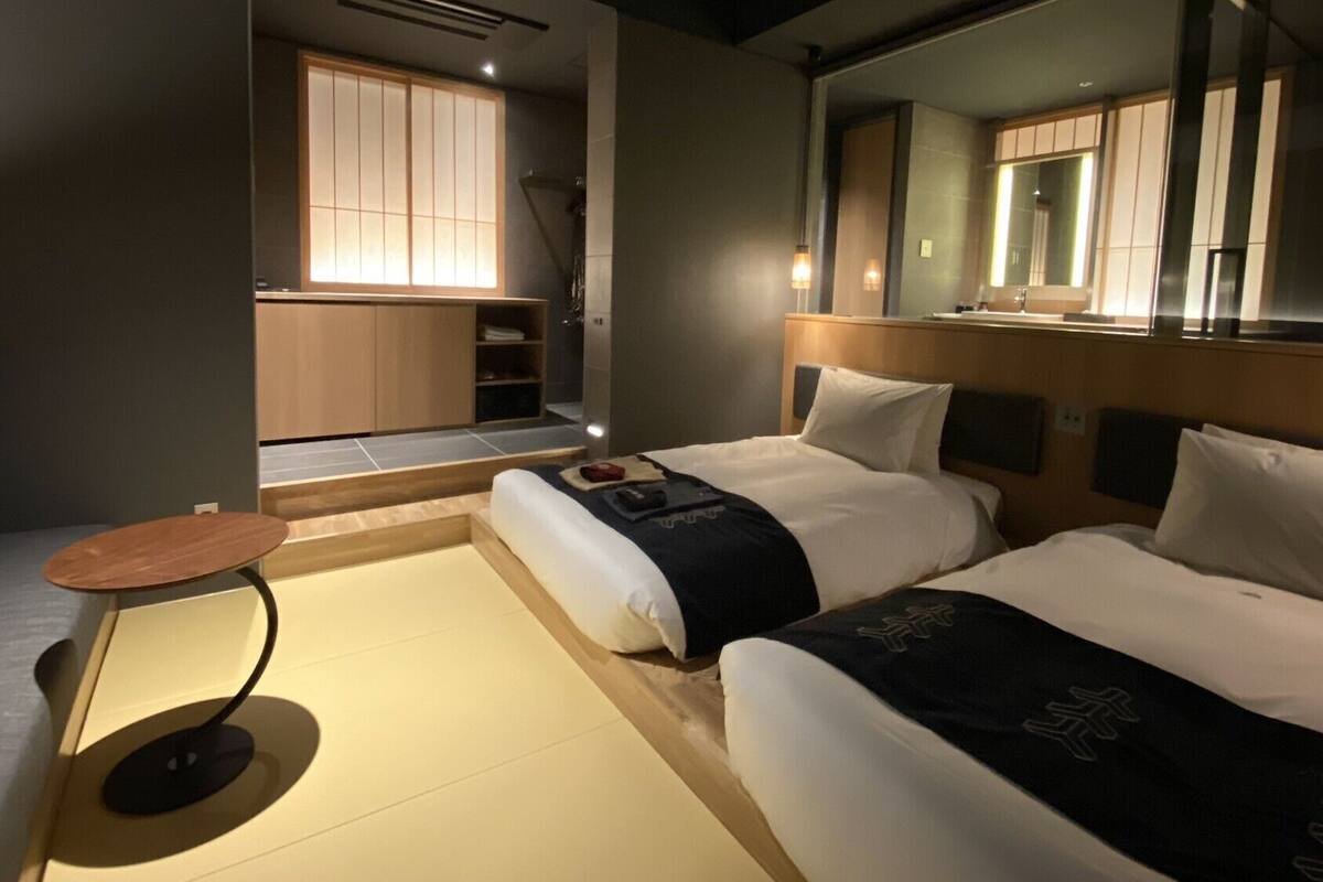 Quarto de hotel com duas camas, uma mesinha, um espelho e vista para um tradicional portas corrediças japonesas.