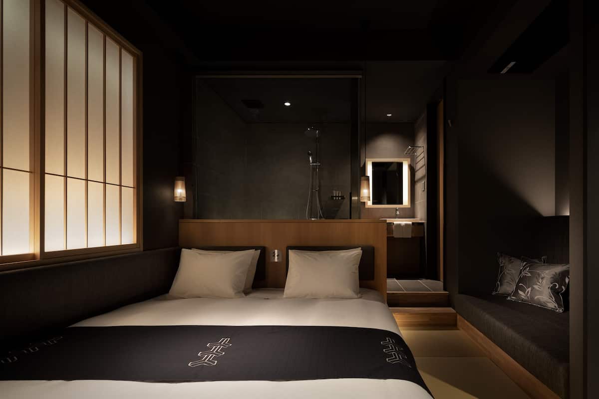 Quarto de hotel moderno com uma cama grande, cabeceira de madeira, banheiro pequeno com chuveiro e vista para a cidade.