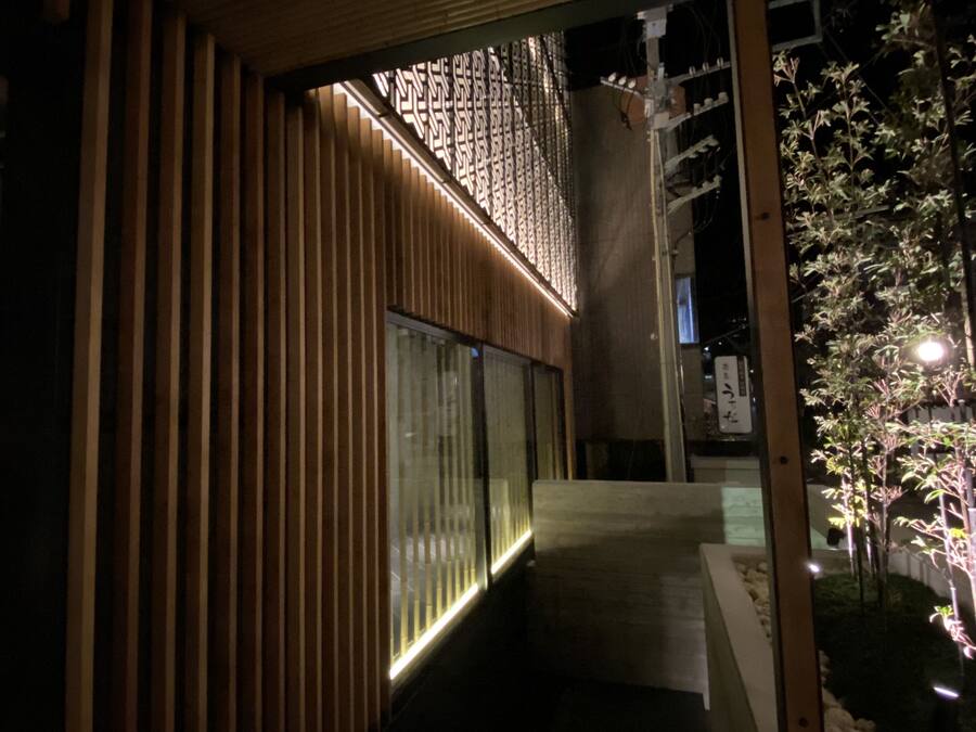 Um edifício moderno com ripas verticais de madeira e painéis iluminados, uma porta de vidro e uma pequena árvore com luzes.