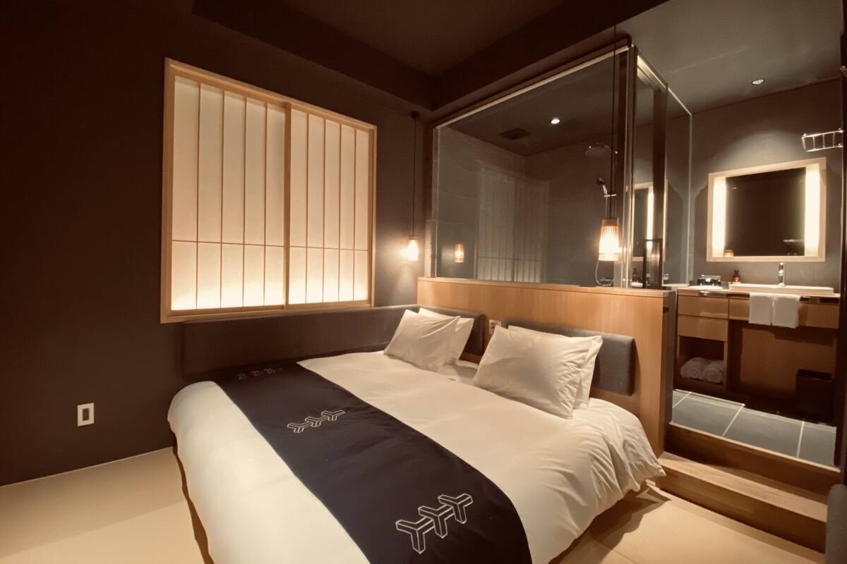Quarto de hotel moderno com uma cama grande, banheiro com espelho e janela com persianas.