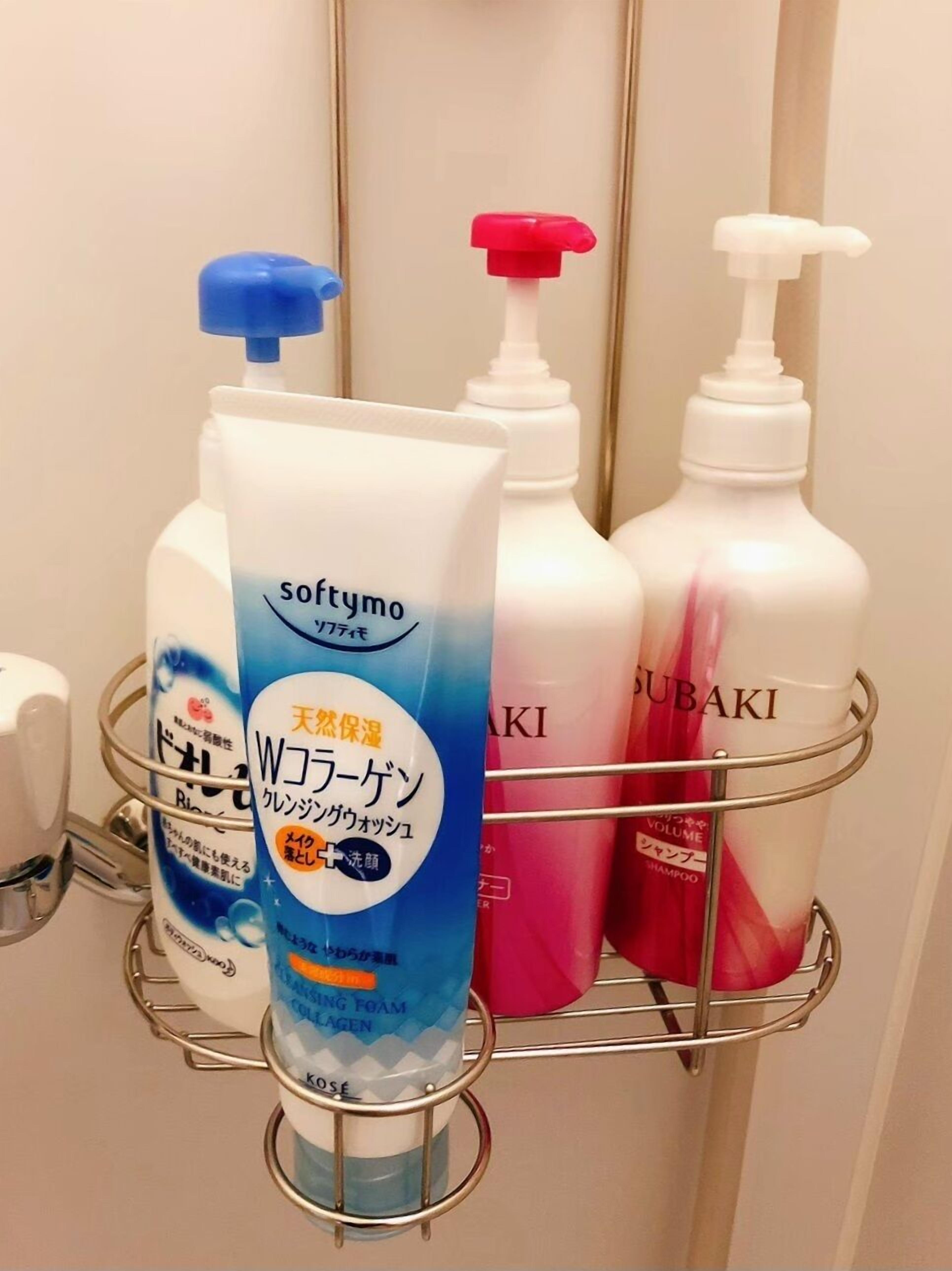 Uma prateleira no banheiro com diversos produtos de higiene pessoal, incluindo uma garrafa de shampoo Softymo, uma garrafa de shampoo Subaki e uma embalagem de espuma.