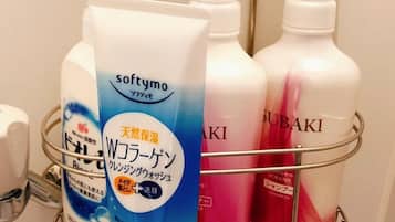 Uma prateleira no banheiro com diversos produtos de higiene pessoal, incluindo uma garrafa de shampoo Softymo, uma garrafa de shampoo Subaki e uma embalagem de espuma.