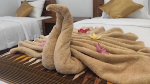 Quarto de hotel com duas camas, cada uma com roupa de cama branca e almofadas douradas. Uma toalha marrom está pendurada sobre a cama em primeiro plano, e há flores rosa e amarelas sobre a cama.