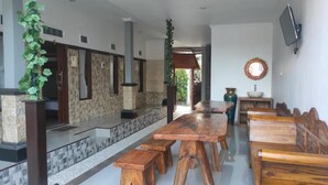 Um espaço amplo e coberto com uma longa mesa de madeira e bancos, piso ladrilhado e televisão fixada na parede.
