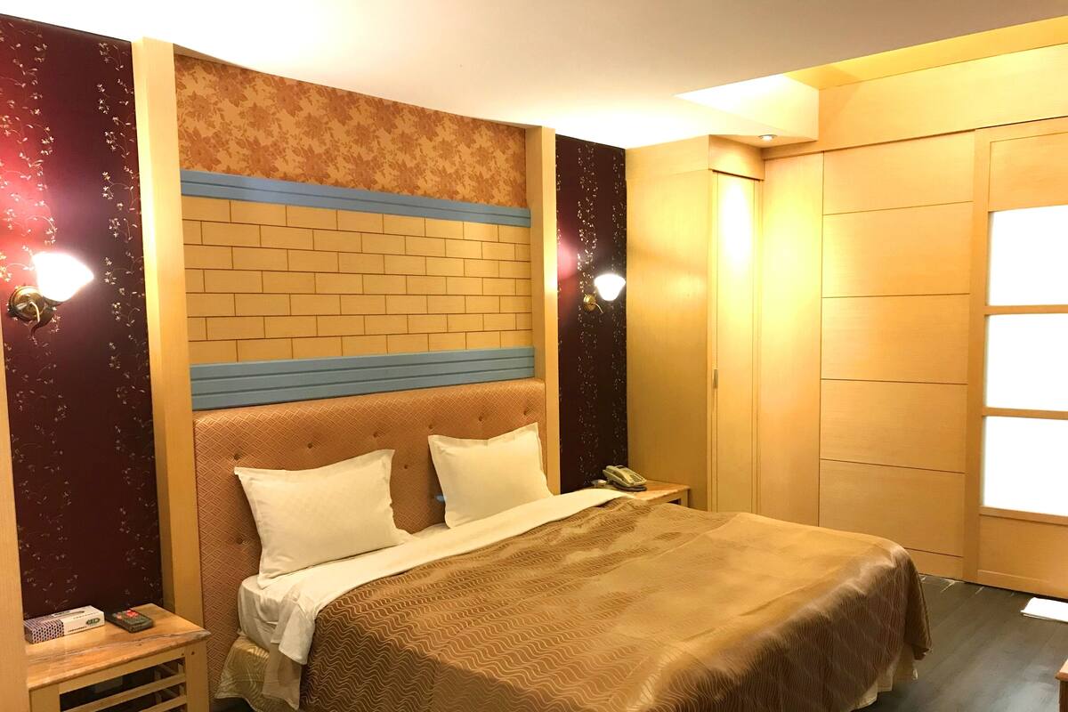Quarto de hotel com cabeceira de tijolos, mesa de cabeceira de madeira, mesa lateral com telefone e janela com persianas.