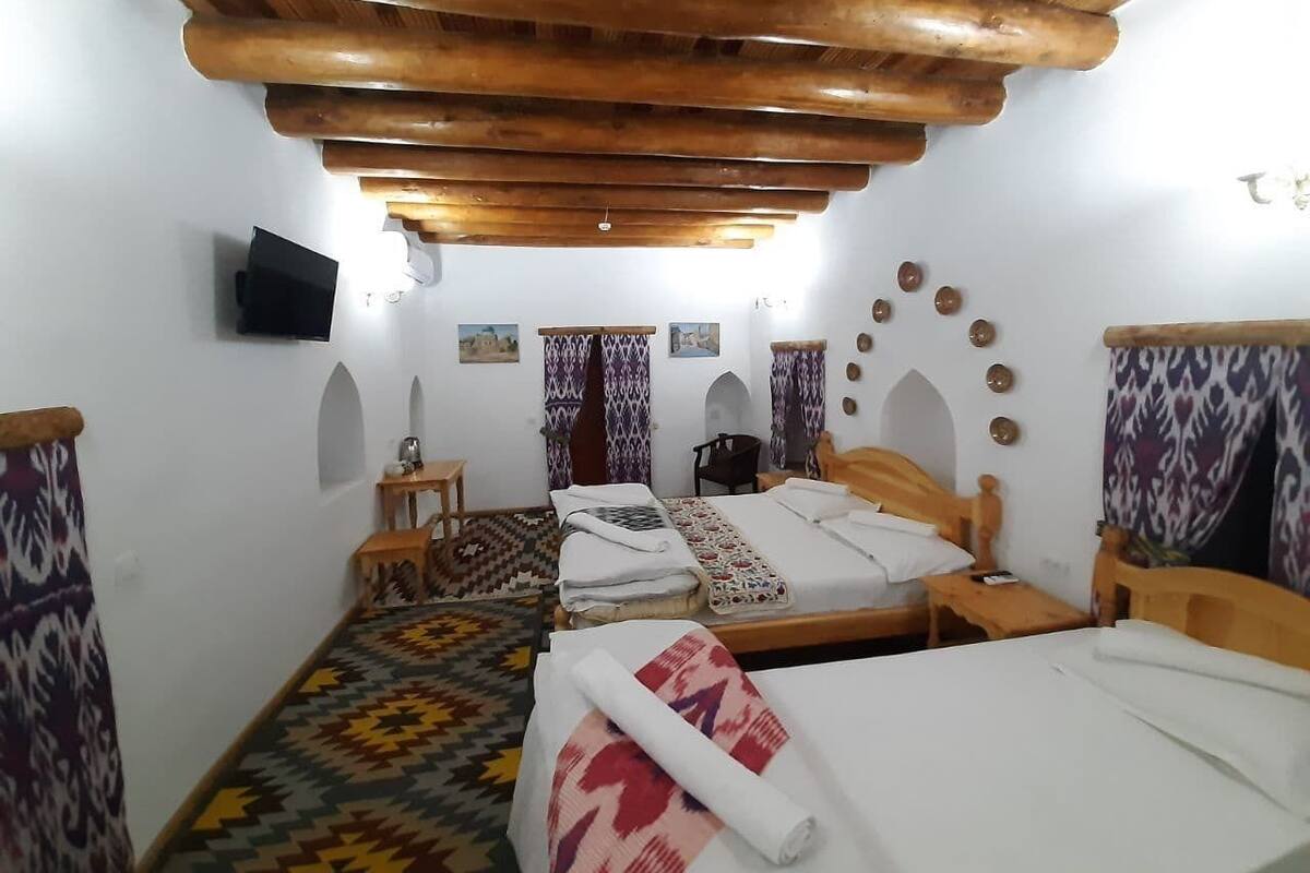 Quarto com teto de madeira, cama com colcha estampada, uma mesinha e uma televisão fixada na parede.