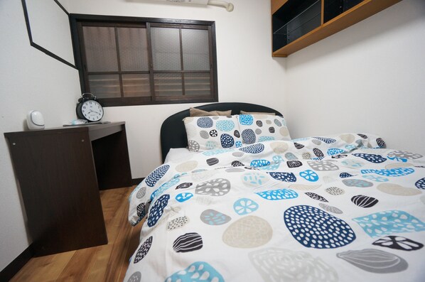 Quarto com cama, mesa de cabeceira, relógio de parede e janela com persianas.