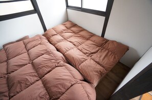 Uma cama de solteiro com colcha rosa num quarto com paredes brancas e chão de madeira.
