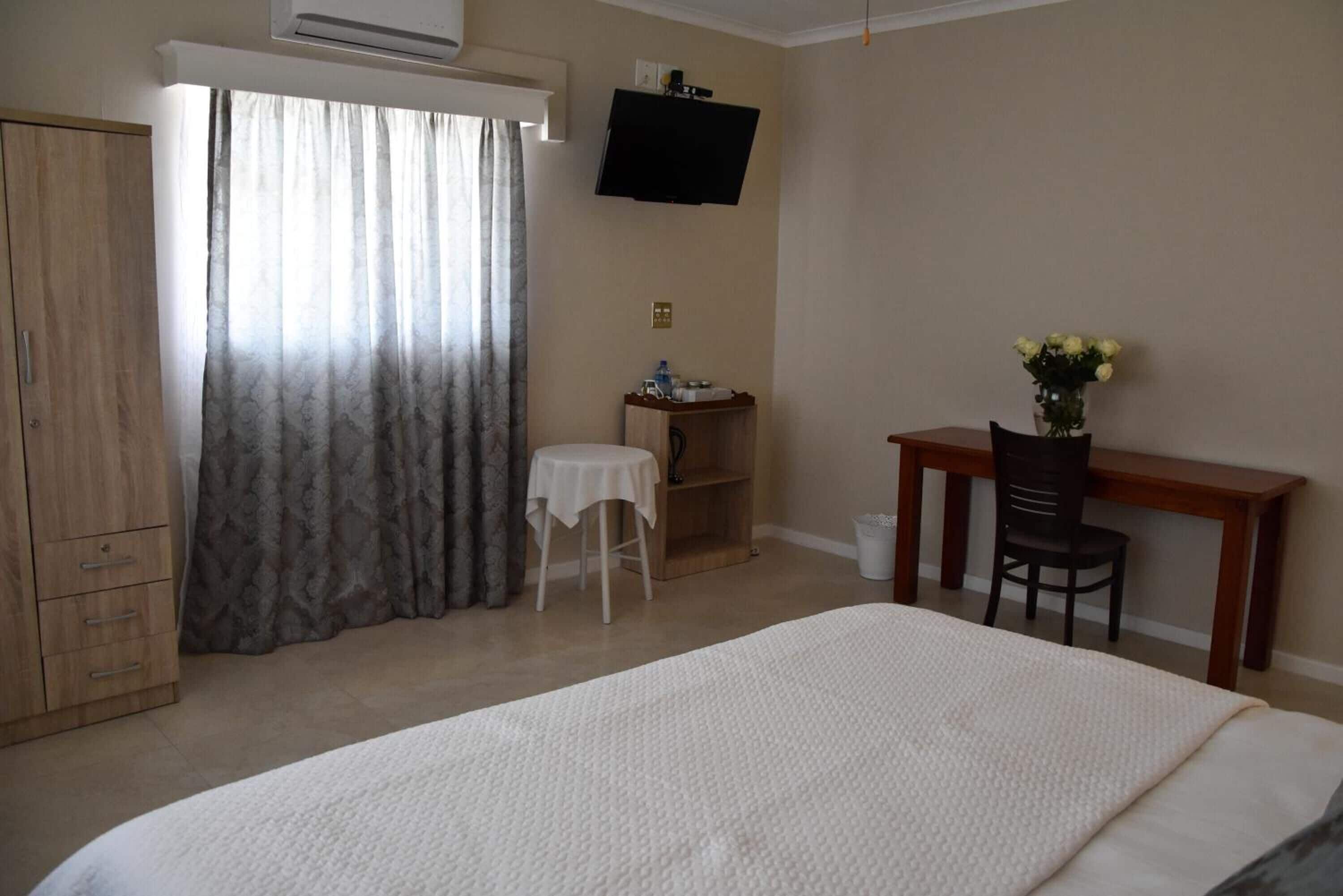 Quarto de hotel com cama, escrivaninha, cadeira, mesinha, guarda-roupa e televisão presa à parede.