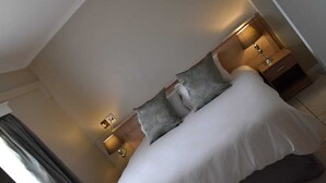 Uma cama bem arrumada com roupa de cama branca e cinza, duas mesas de cabeceira com luminárias e uma janela com cortinas.