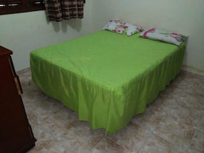 Quarto com cama coberta por um edredom verde, guarda-roupa de madeira e janela com cortinas.