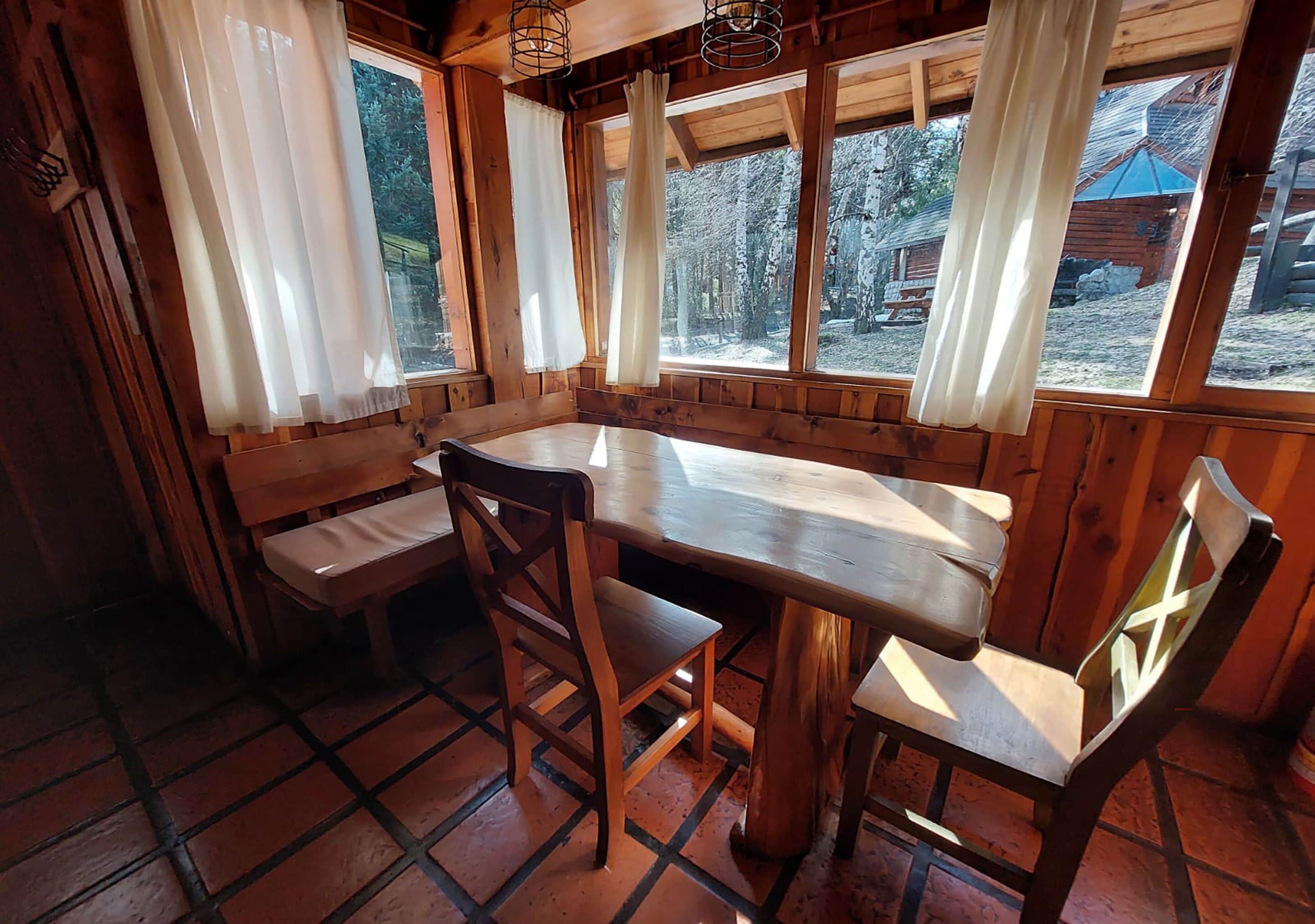 Interior de cabana de madeira com mesa central, duas bancos e janelas amplas com cortinas.