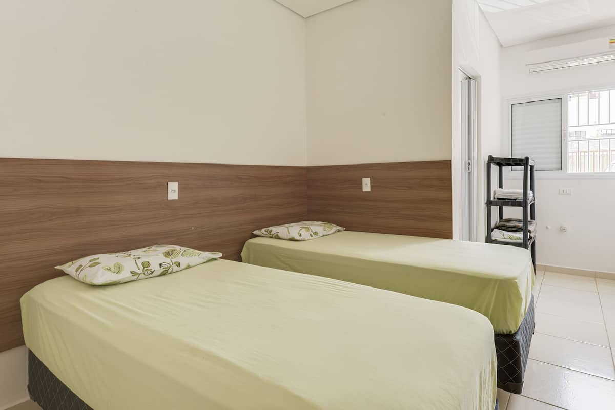 Quarto com duas camas de solteiro, cabeceira de madeira, janela com persianas, uma cadeira e uma porta.