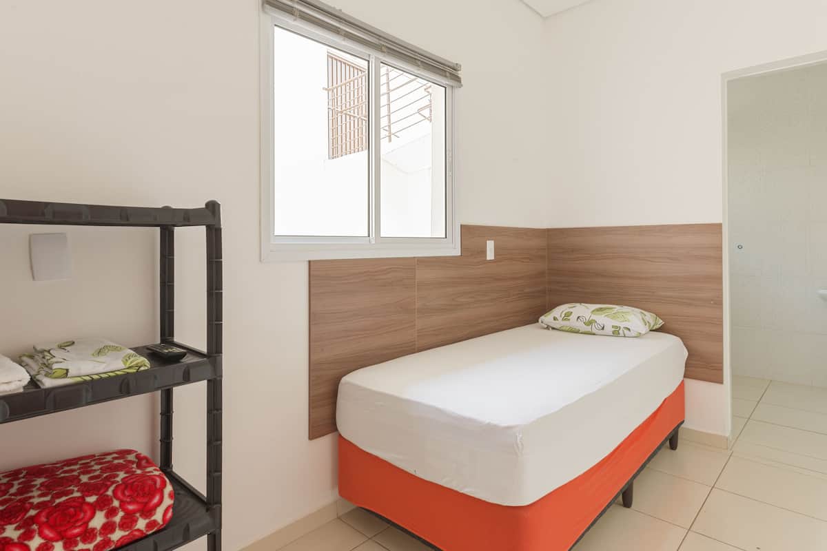 Quarto pequeno com uma cama de solteiro, cabeceira de madeira, janela com grades de metal, beliche com almofada estampada em vermelho e branco, e piso de azulejos.