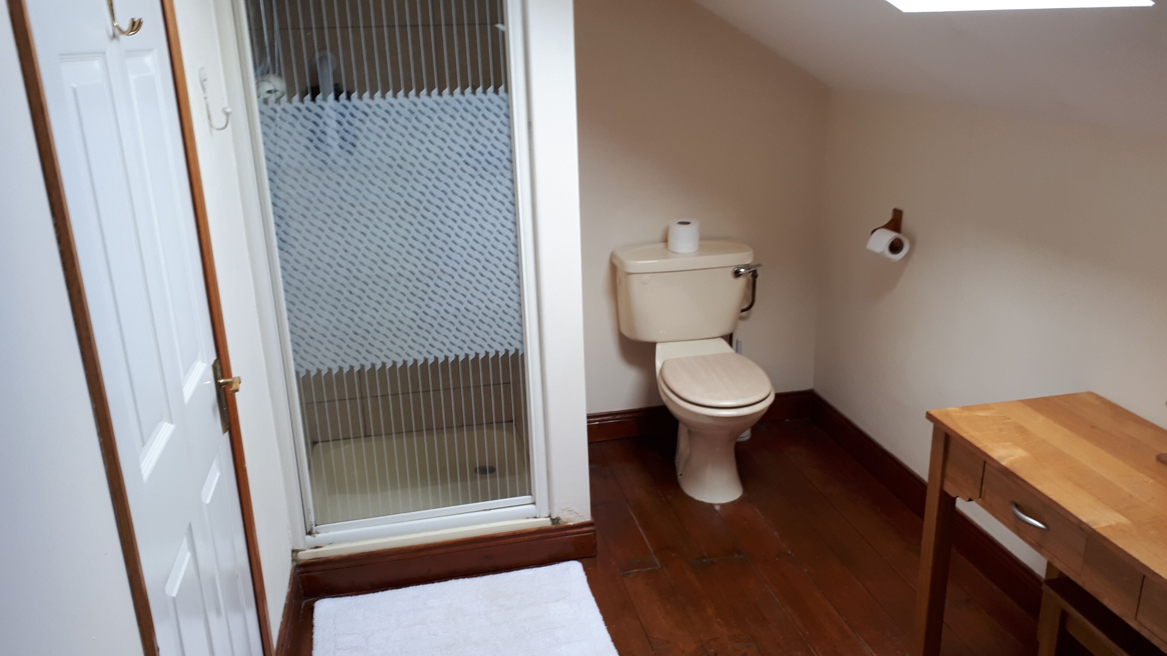 Casa de banho pequena com sanita, um banco de madeira e zona de duche com porta de vidro.
