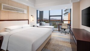 Quarto de hotel com uma cama grande, uma escrivaninha, uma cadeira e uma televisão. Há uma janela com vista para edifícios e colinas.