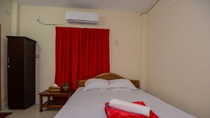 Uma cama de solteiro com roupa de cama branca, um cobertor vermelho e uma cabeceira de madeira. Uma pequena mesa de cabeceira de madeira com uma luminária e uma caixa fica ao lado da cama. Uma cortina vermelha cobre a janela, e um aparelho de ar condicionado está fixado na parede acima da cama.