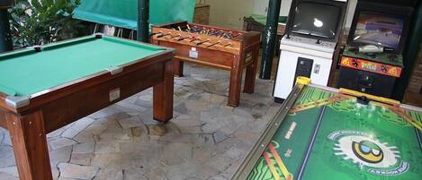 Uma mesa de bilhar, uma mesa de futebol de mesa e um fliperama em um espaço com piso de pedra e paredes verdes.