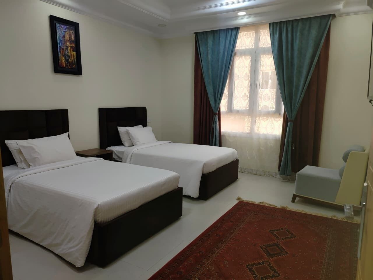 Quarto de hotel com duas camas, um tapete vermelho, uma cadeira e um quadro na parede.